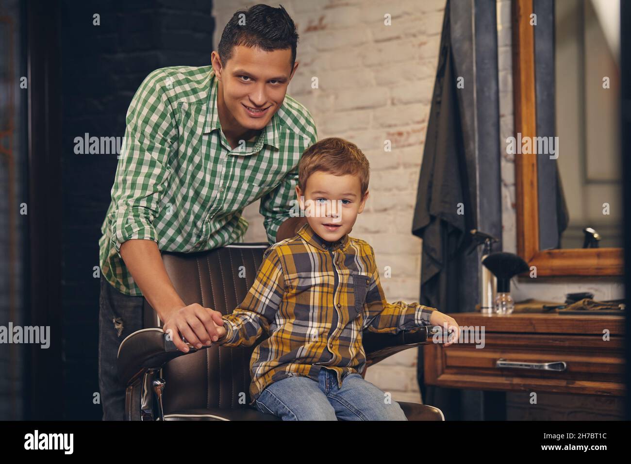 stylish little kid sitting on chair at barbershop with his young father ...