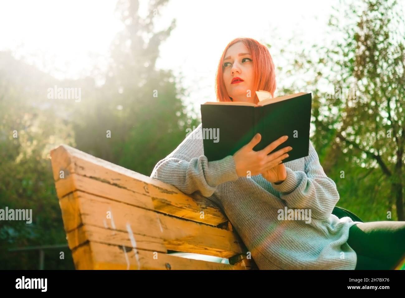 Adult girl is sitting on bench and reading book on nature in park ...