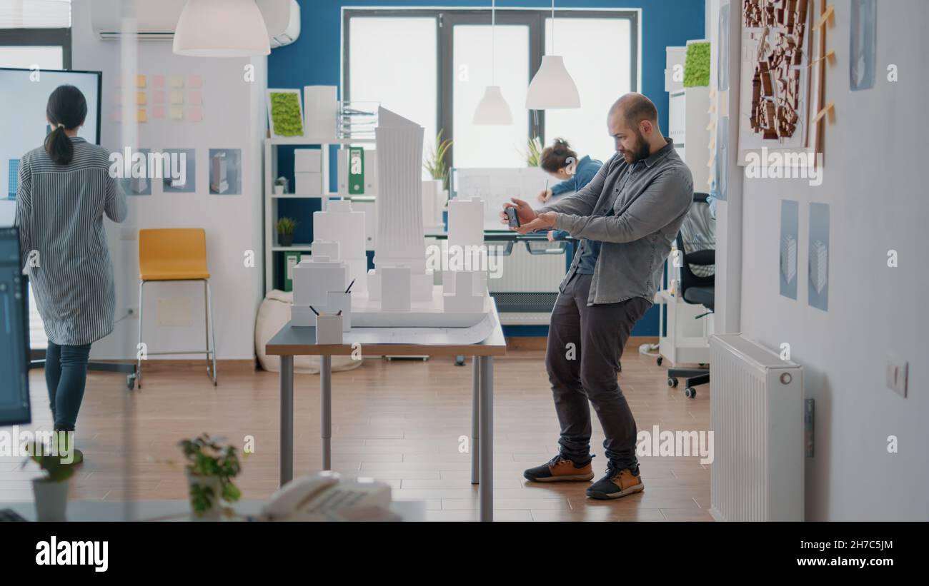 Architect Using Mobile Phone To Create Blueprints Plan With Building Model And Maquette For