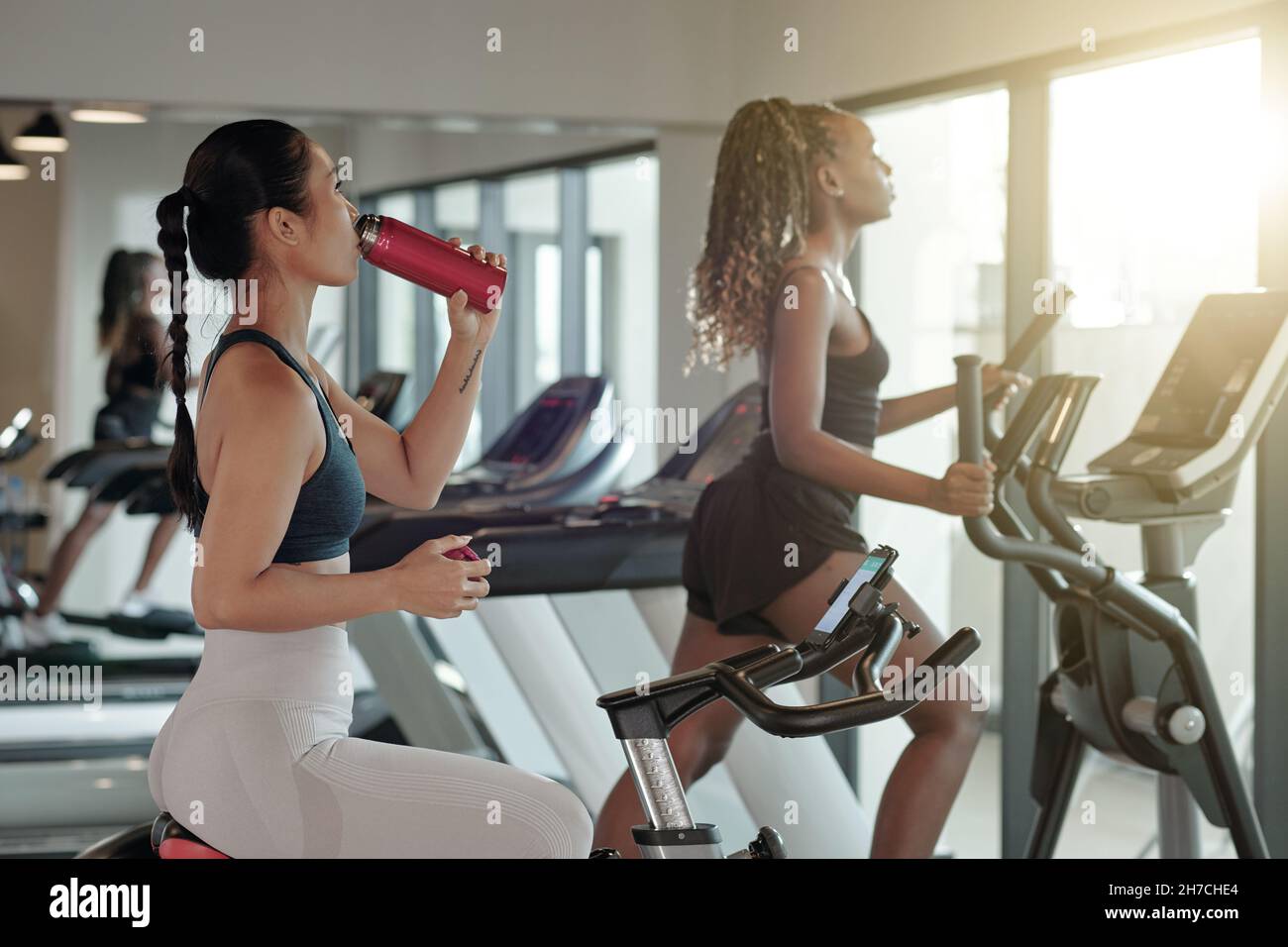 Beautiful thirsty fit young woman sitting on exercise bike and drinking ...