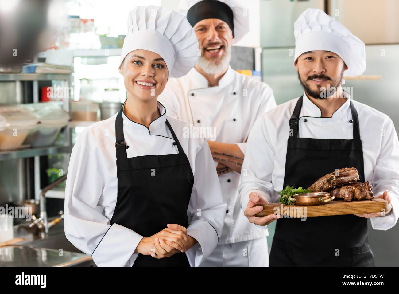 Asian chef holding roasted meat near cheerful colleagues in kitchen ...