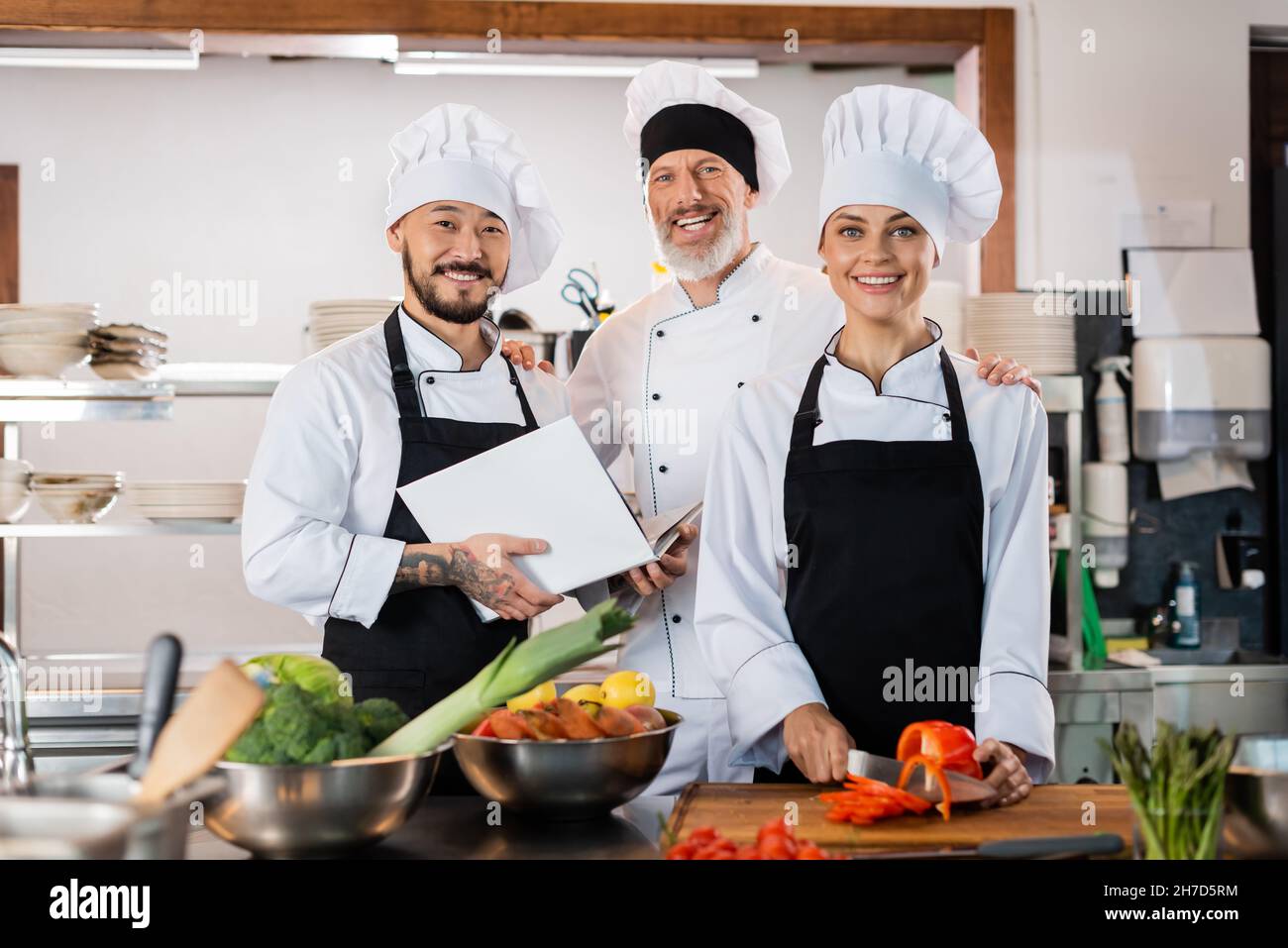 Asian chef holding cookbook near smiling colleagues and vegetables in ...