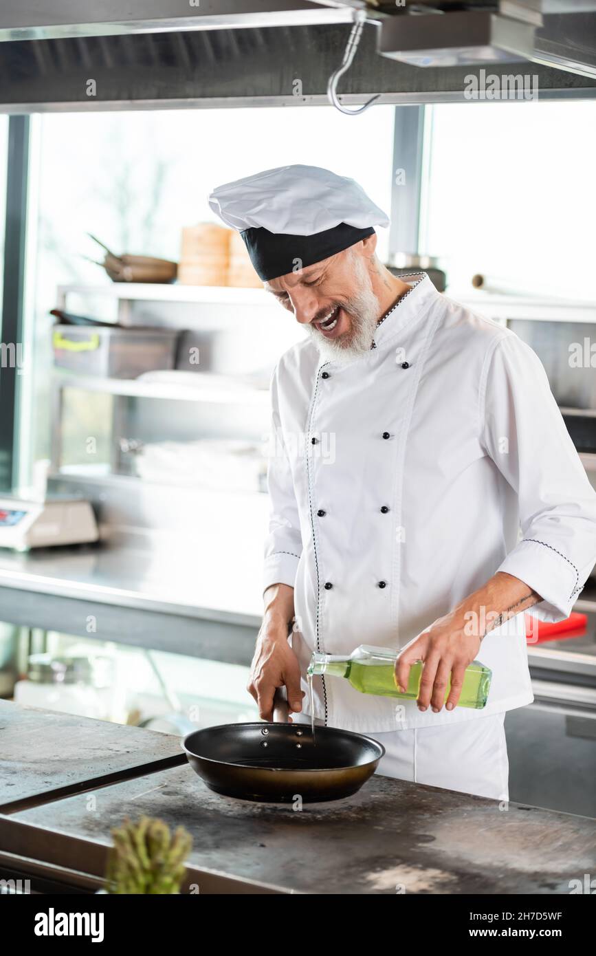 Positive mature chef pouring olive oil on frying pan on cooktop in ...