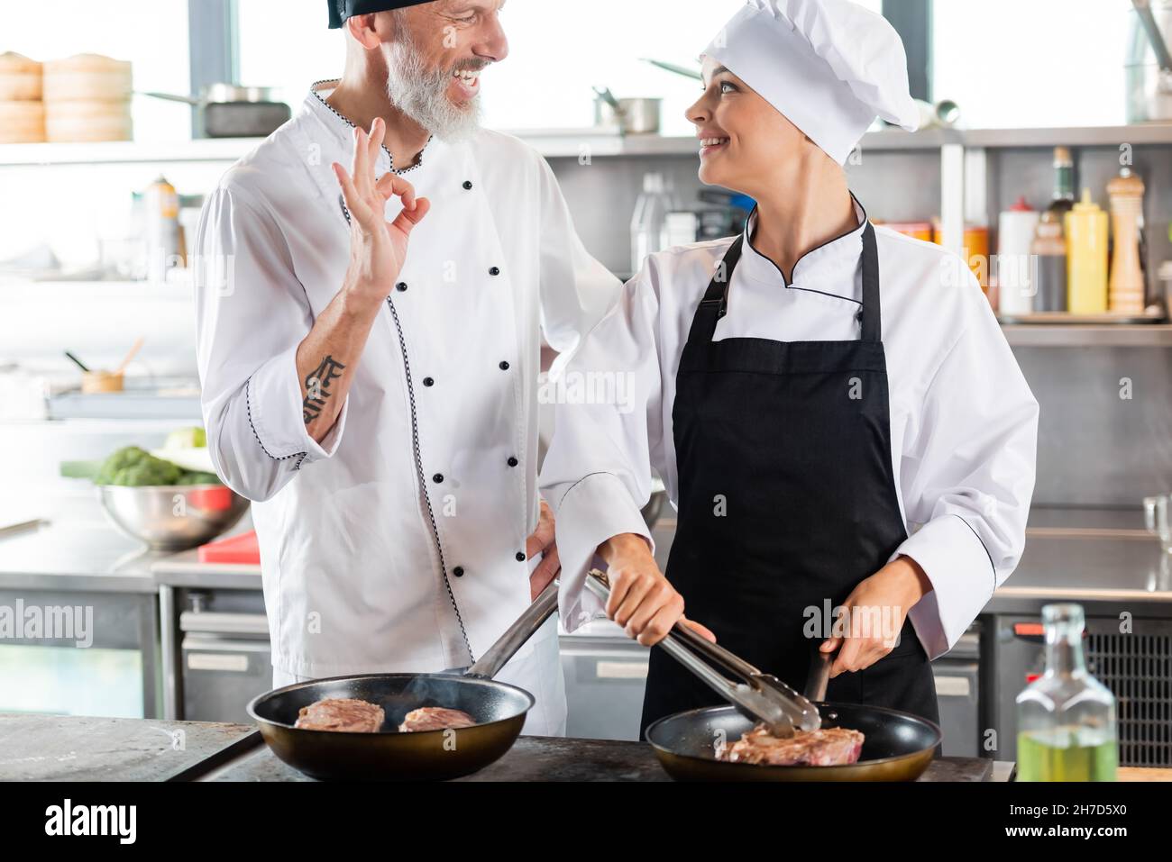 Smiling chef showing ok gesture near colleague cooking meat in kitchen ...