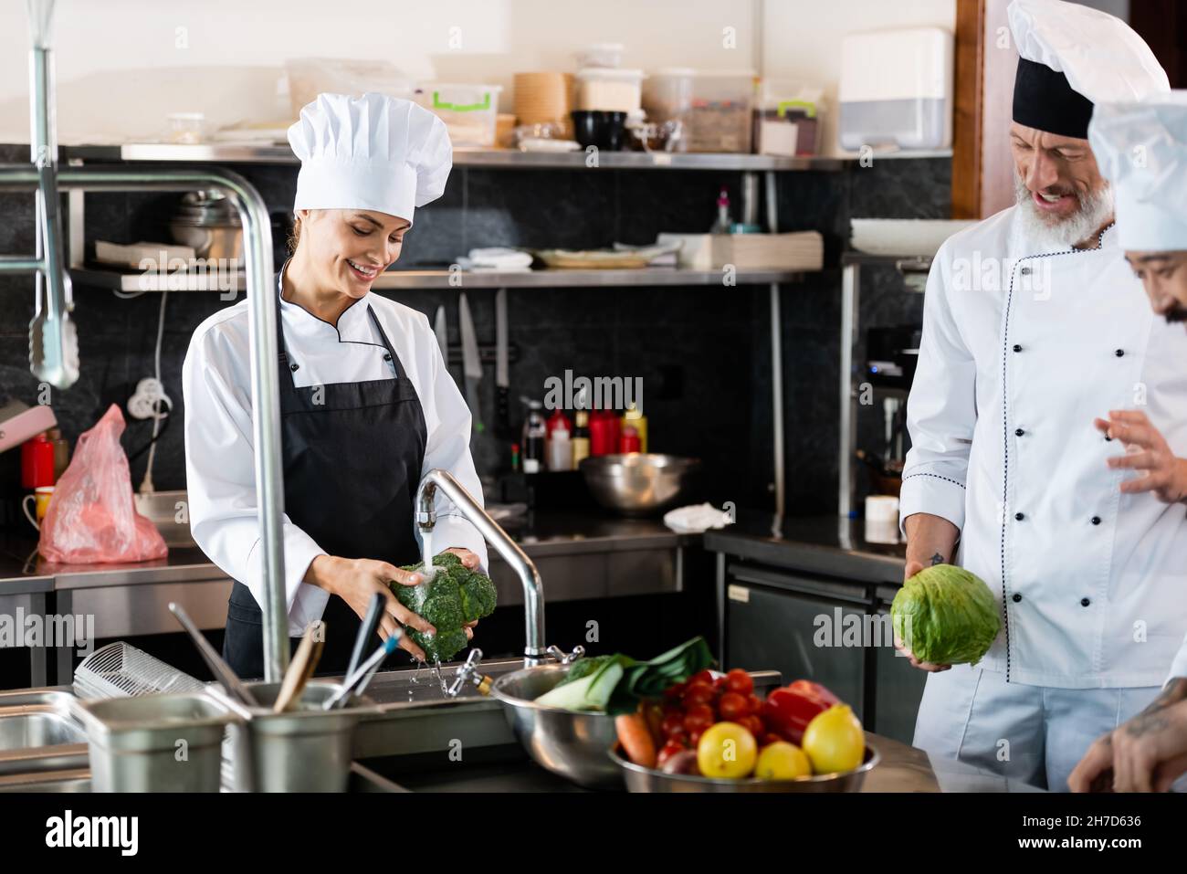 Smiling chef washing broccoli near interracial colleagues talking in ...