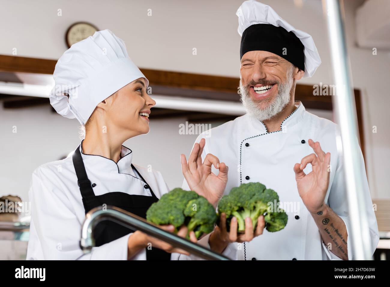 Smiling chef showing ok gesture near colleague with broccoli in kitchen ...