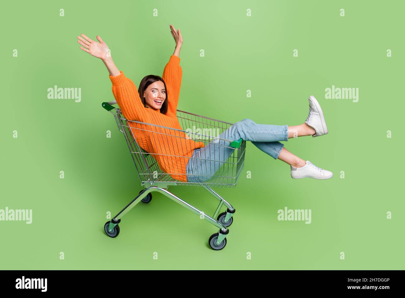 Portrait of attractive cheerful childish girl riding cart having fun isolated over bright green ...