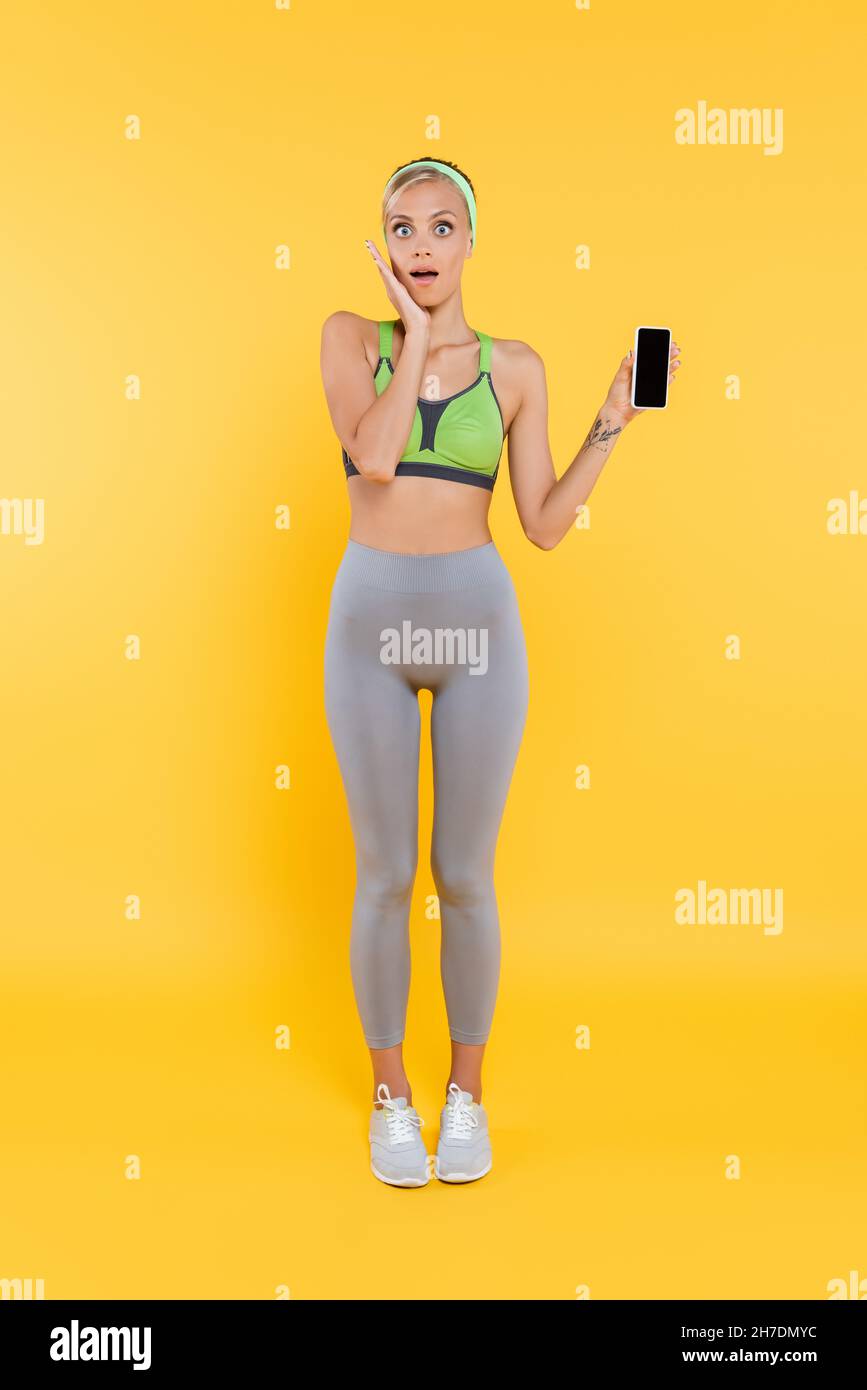 full length view of amazed woman in sportswear touching face while ...