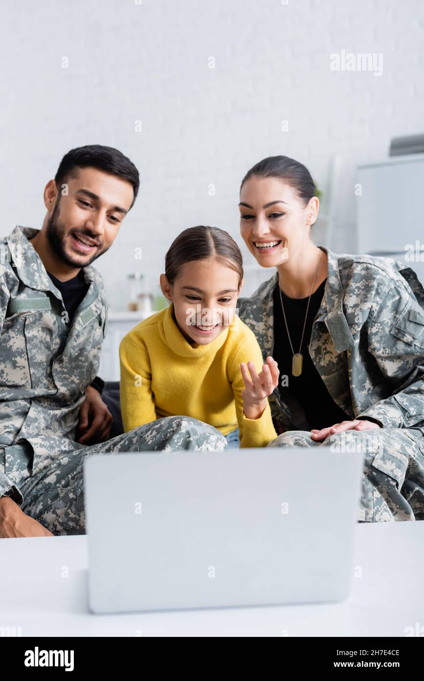 Smiling kid looking at blurred laptop near positive parents in military ...