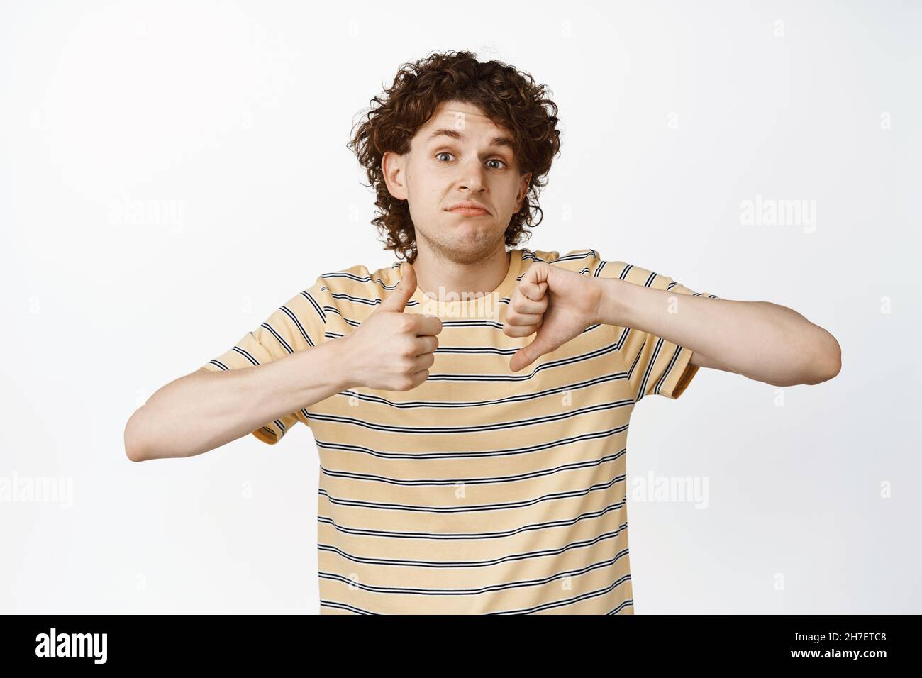 Indecisive young man showing thumbs up and thumbs down, looking ...