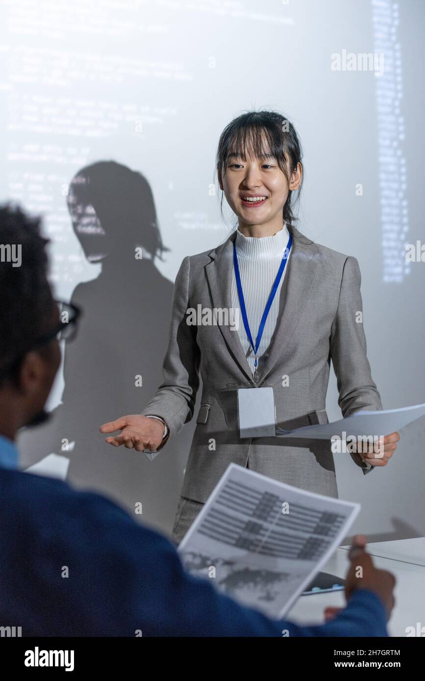 Happy young Asian female with papers standing by interactive whiteboard ...