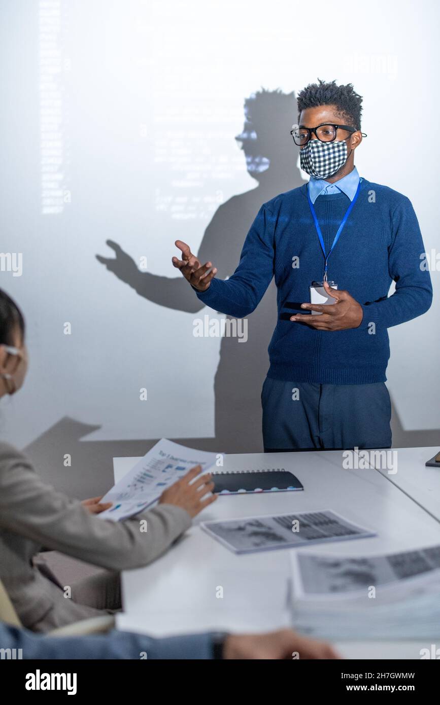 Young African programmer in protective mask standing by interactive ...