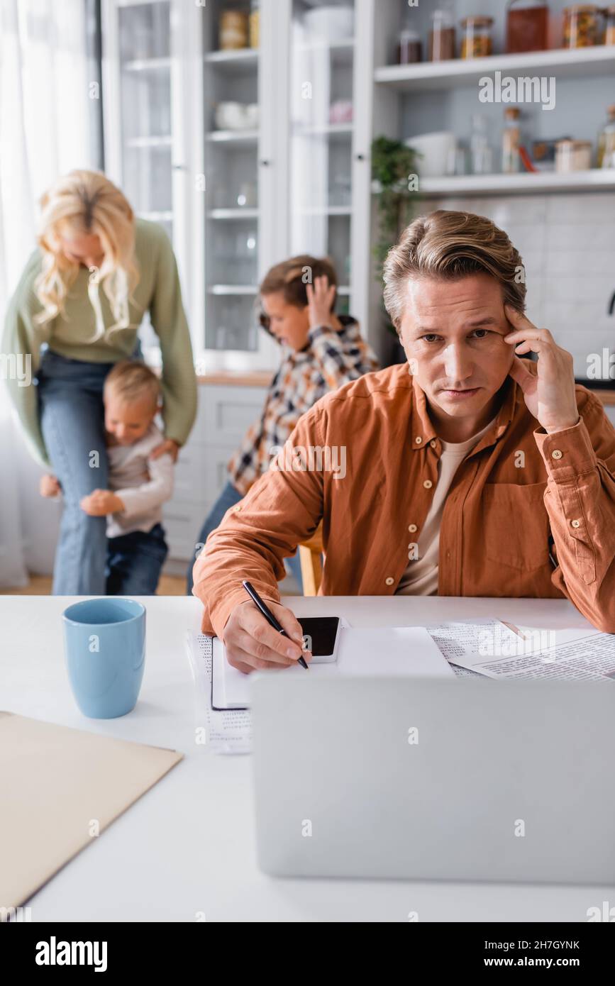 thoughtful man writing in notebook while working in kitchen near mother ...