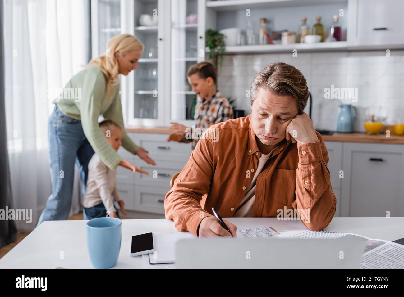tired man working in kitchen while blurred family playing patty cake ...