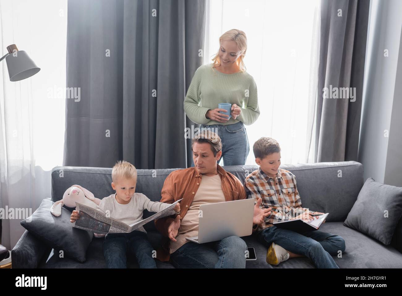 woman with cup of tea standing near husband with laptop and sons with ...