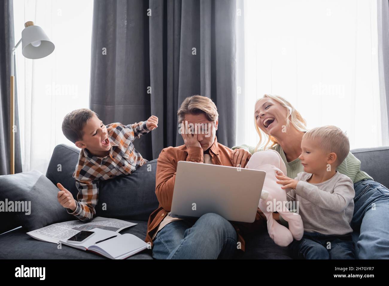 tired man obscuring face with hand while working on laptop near family ...