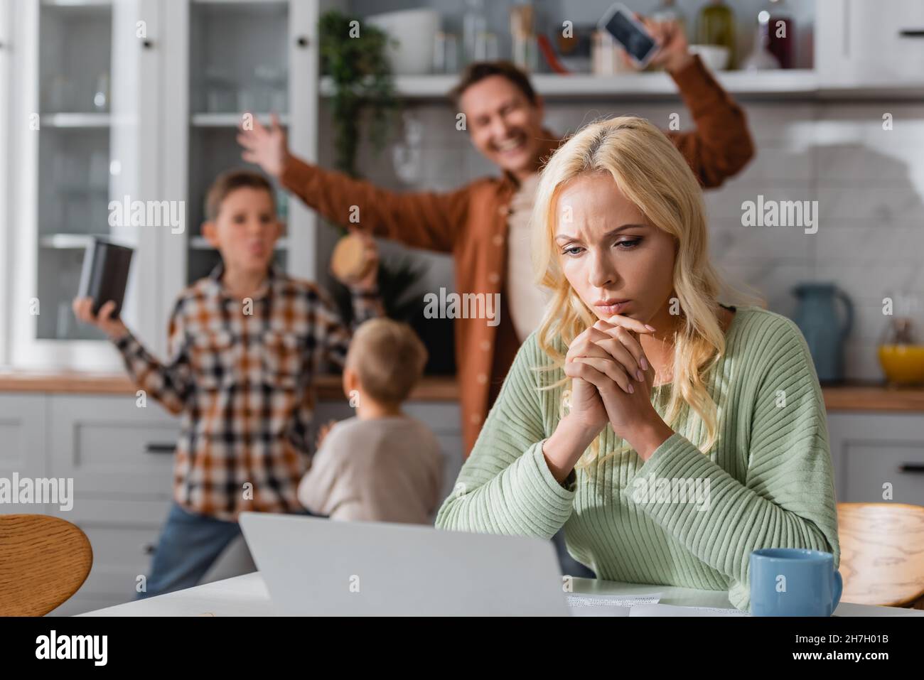 blurred family having fun in kitchen near thoughtful woman working near ...