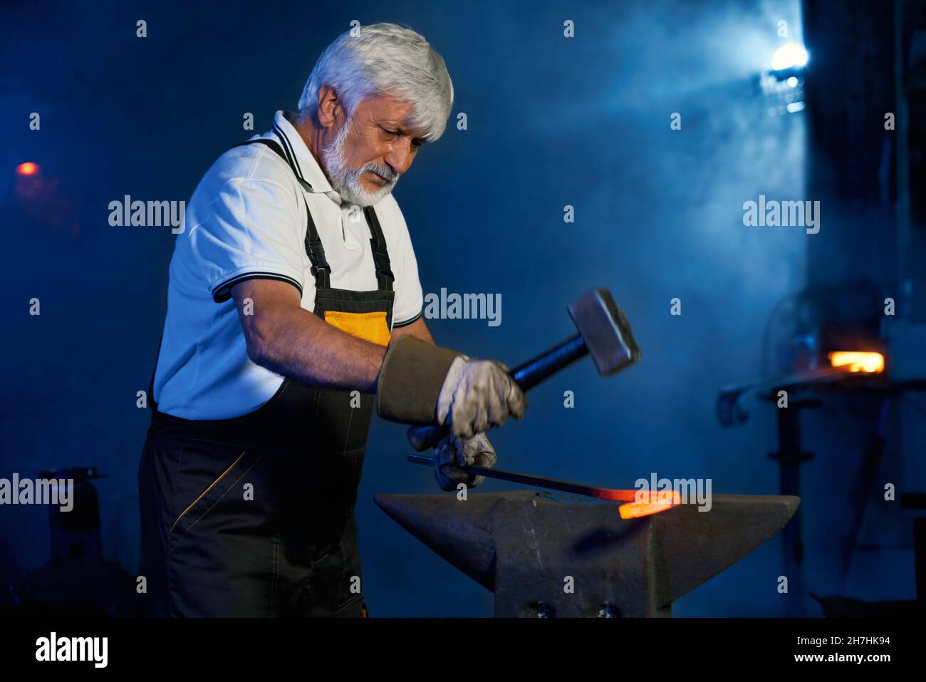 Professional mature blacksmith using heavy hammer for work with molten ...