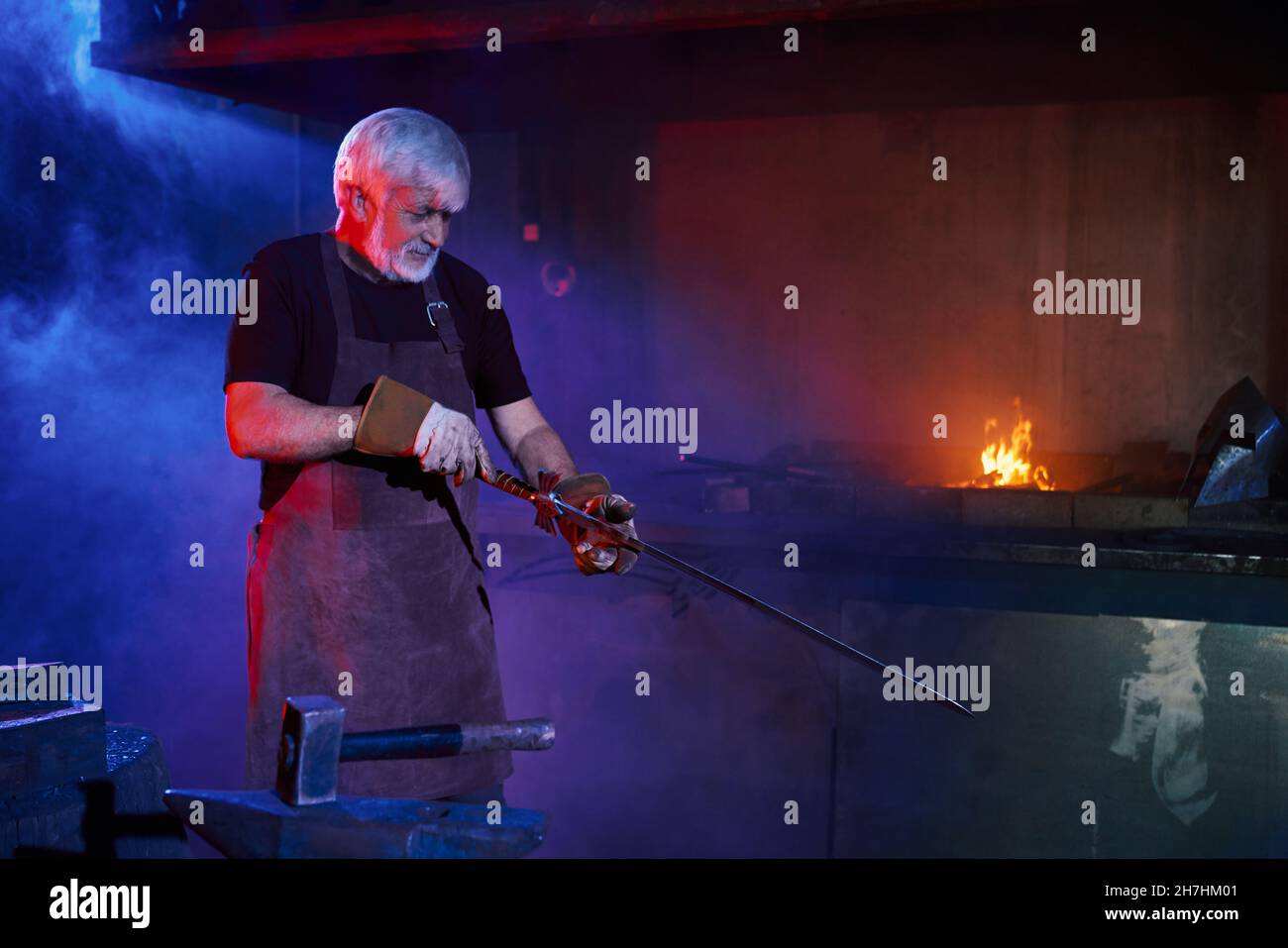Skillful caucasian blacksmith in protective apron and gloves making ...