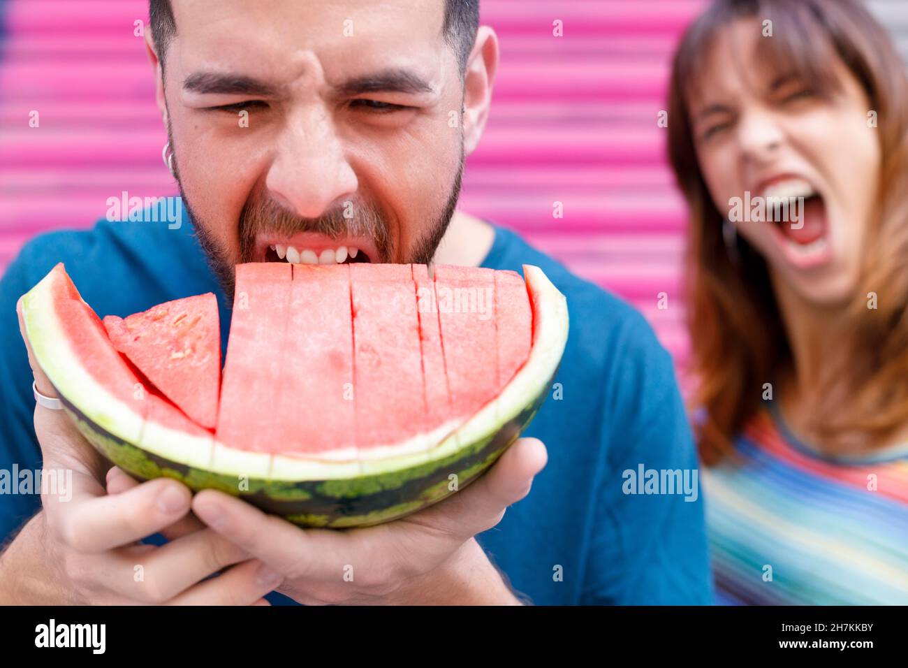 Happy man feeding watermelon to woman in front of pink corrugated wall ...