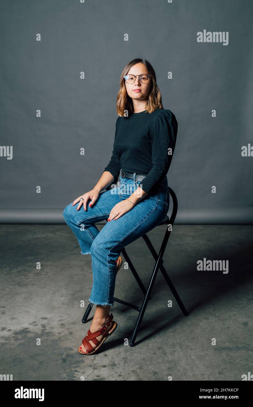 Confident young woman sitting on chair in front of gray backdrop Stock ...