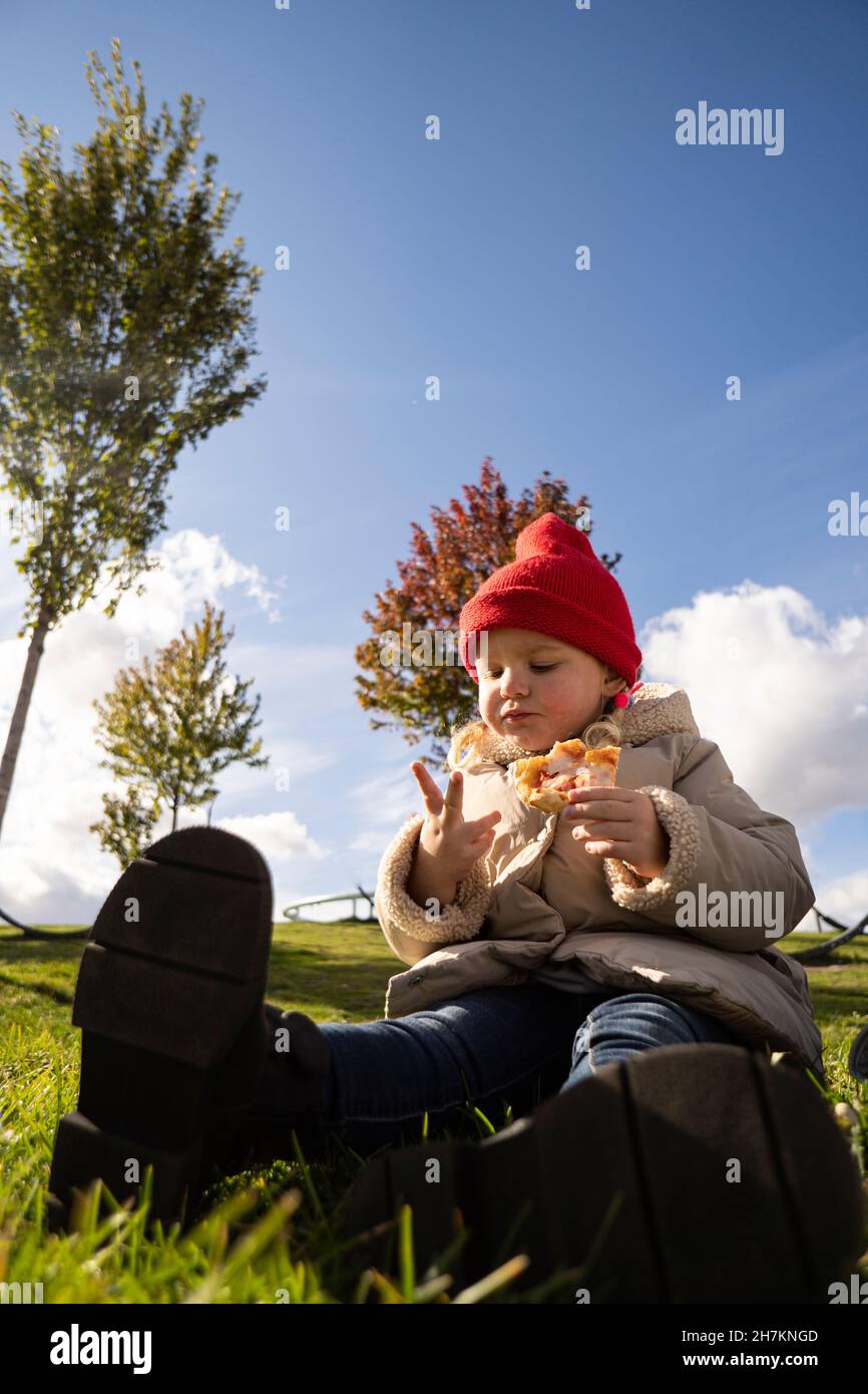 Girl in warm clothing eating pizza while sitting on grass Stock Photo ...