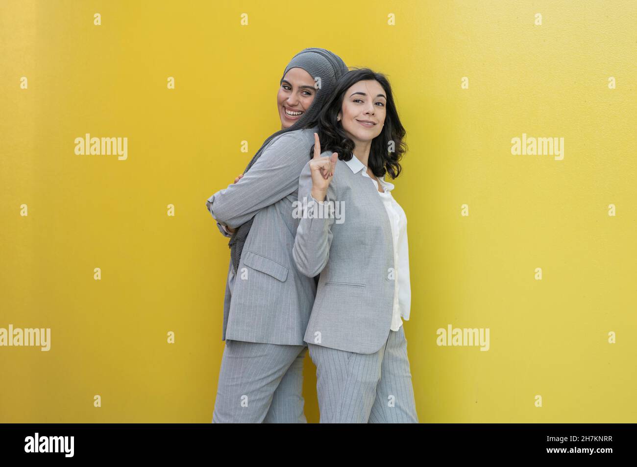 Smiling female colleagues standing back to back by yellow wall Stock ...