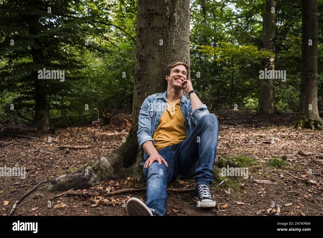 Happy man with hand on chin sitting by tree trunk in forest Stock Photo ...