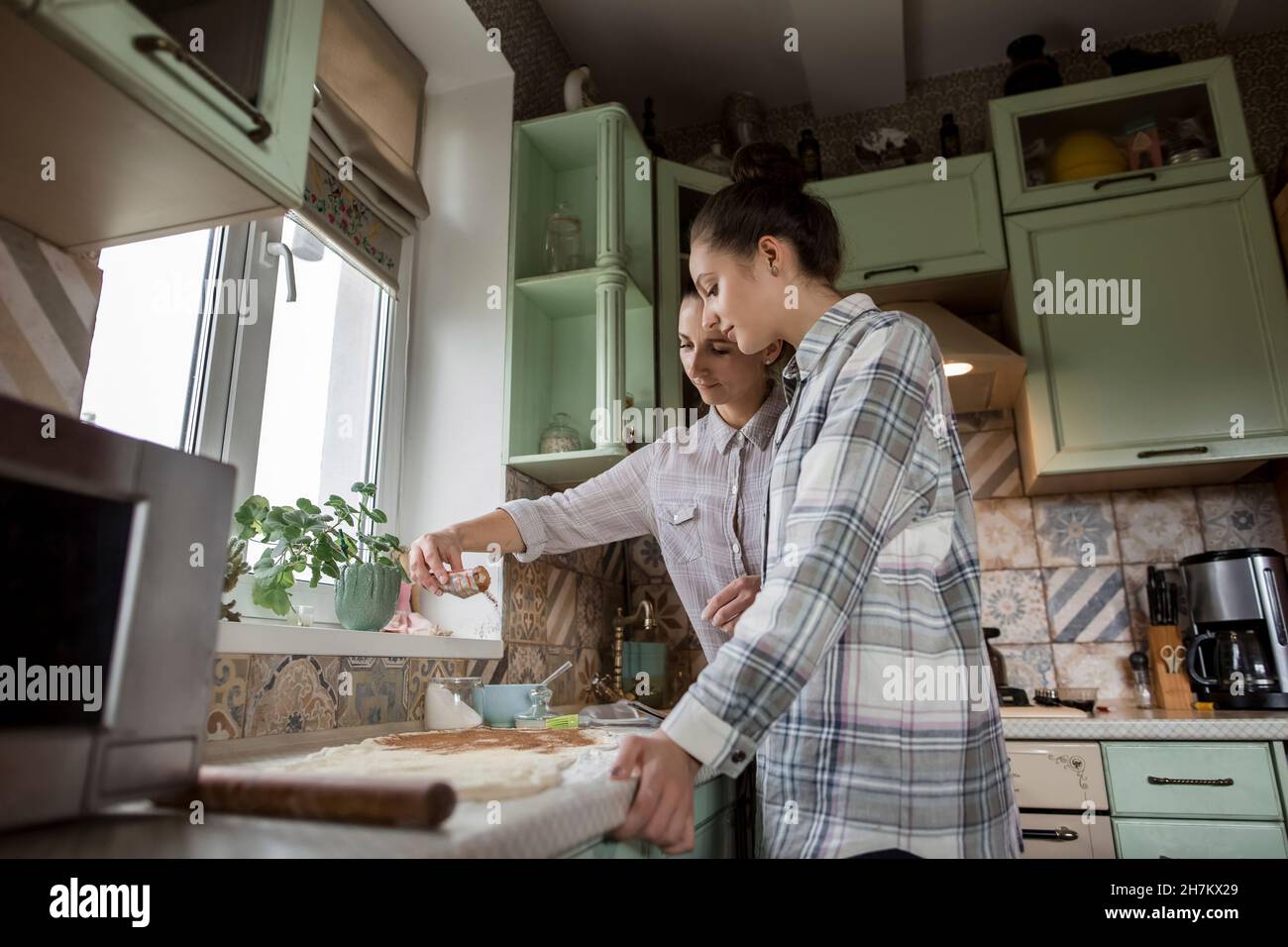 Woman sprinkling cinnamon on dough with daughter at kitchen counter ...
