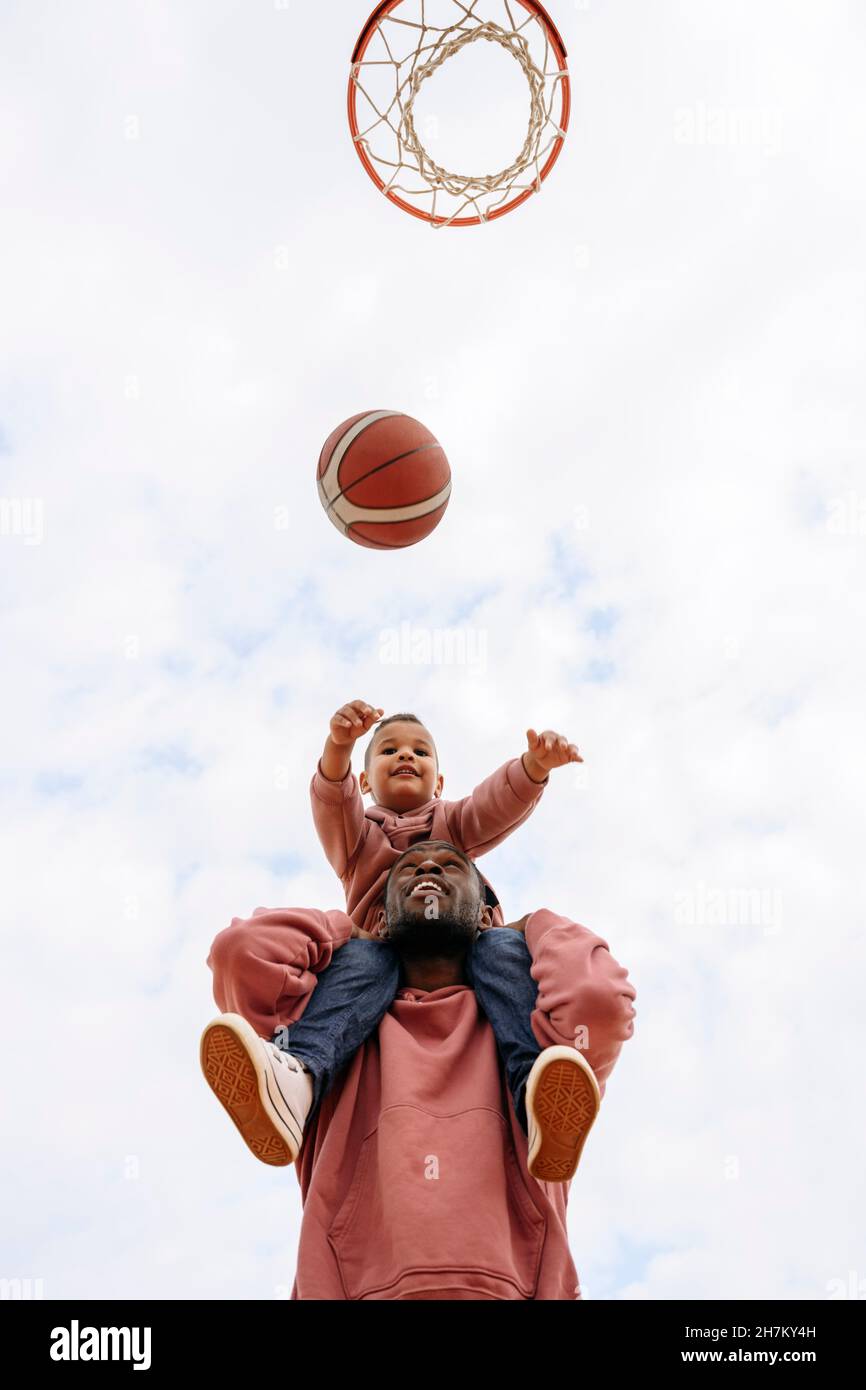 Son throwing basketball towards hoop while sitting on father's ...
