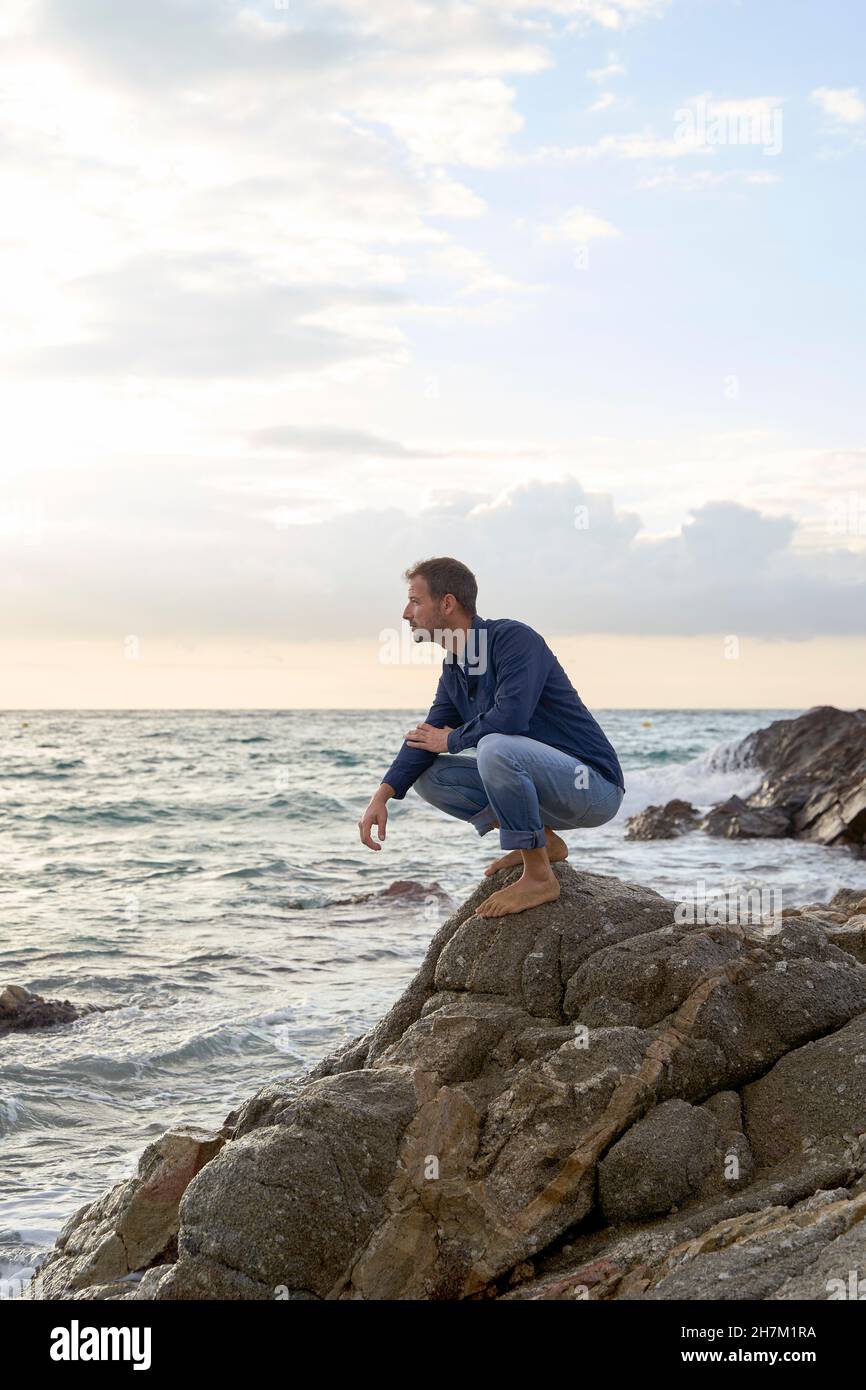 Man crouching on rock by sea at beach Stock Photo - Alamy