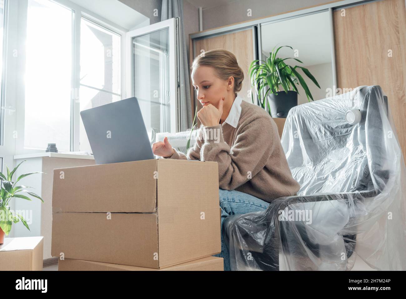 Woman with hand on chin using laptop on cardboard box in new home Stock ...