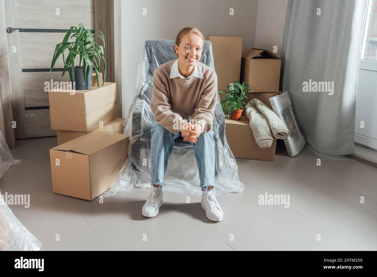 Smiling young woman sitting on armchair wrapped in plastic at home ...