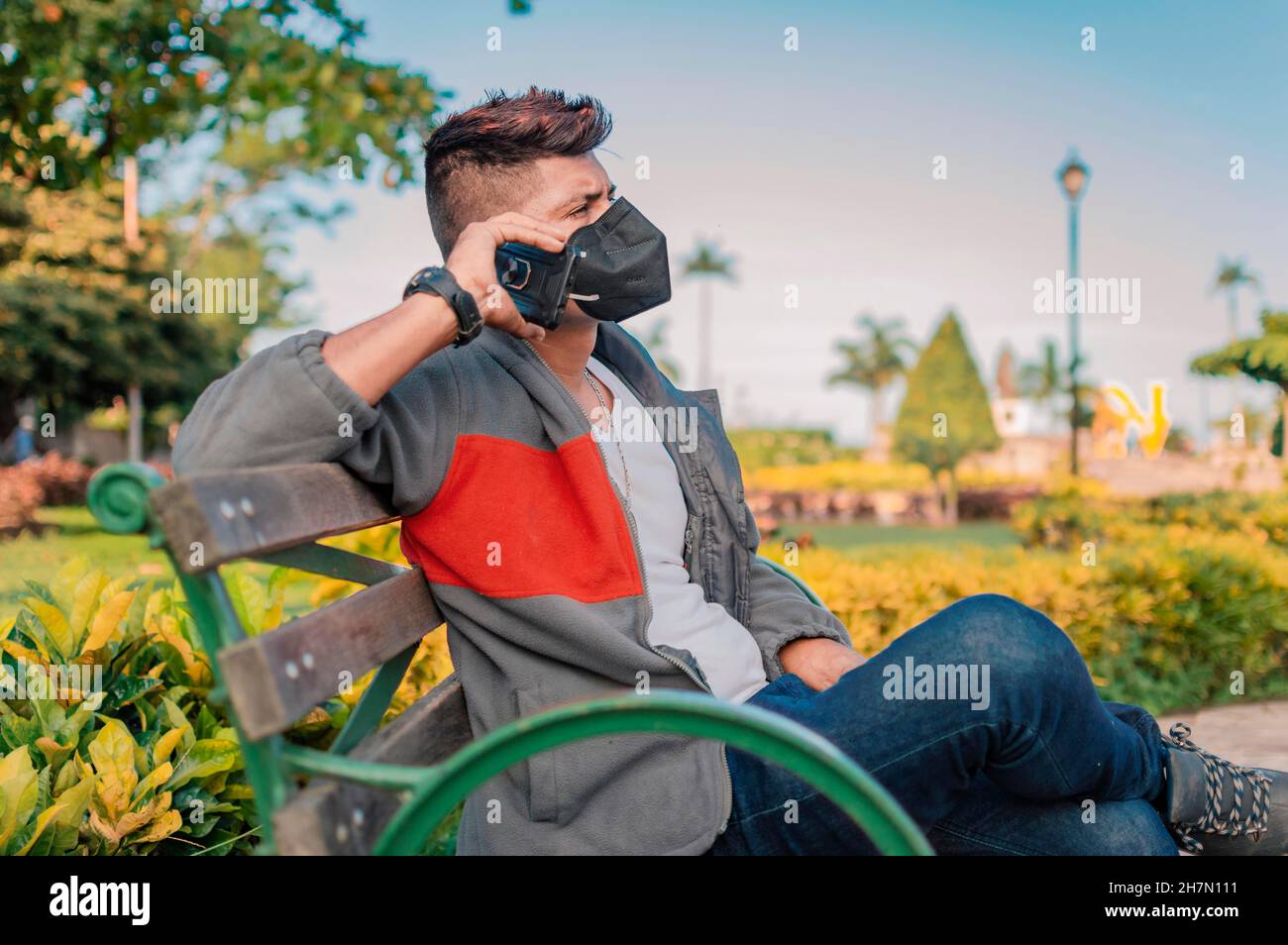 Man sitting on a bench calling on the phone, young man with a mask ...