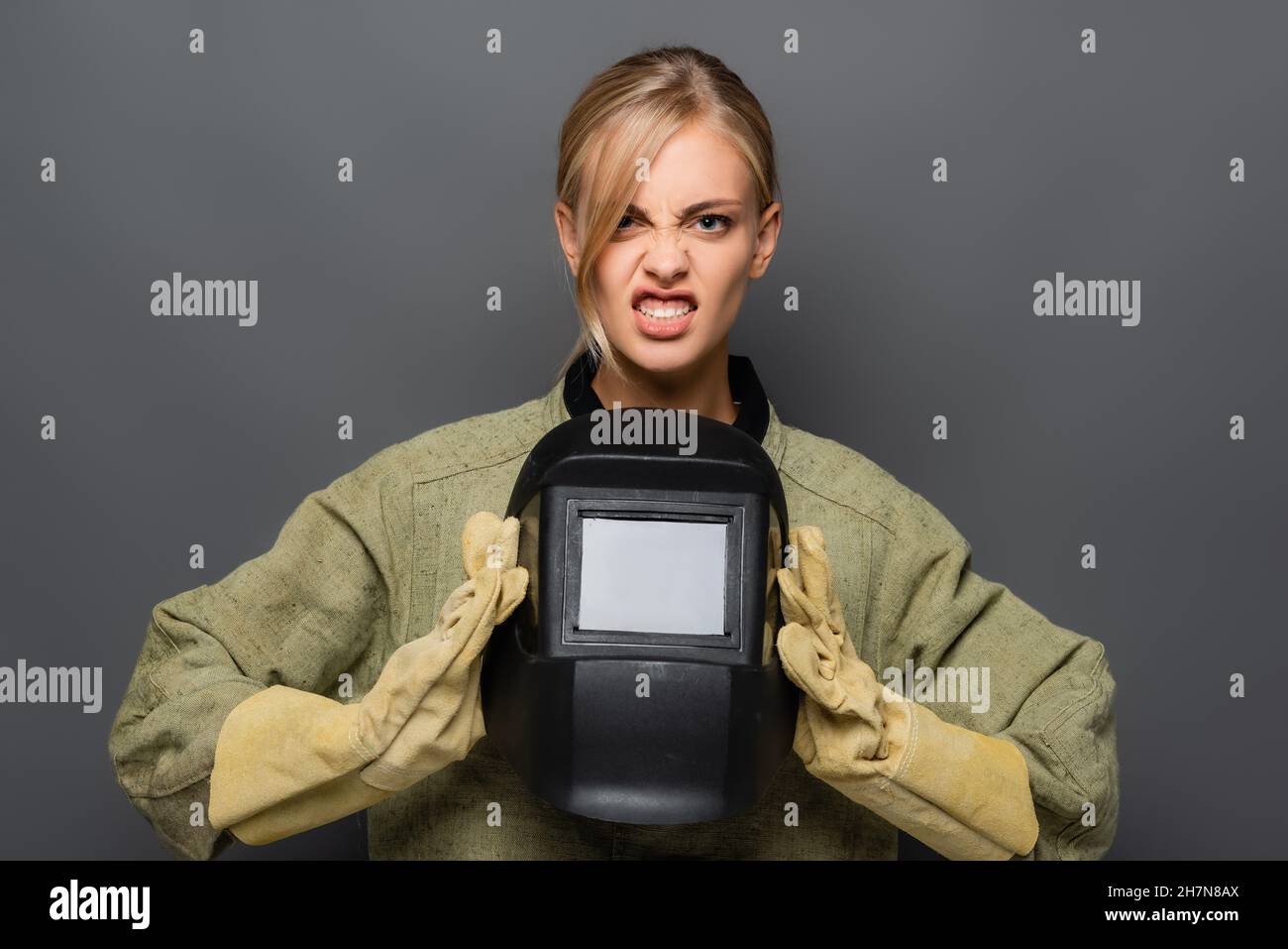 Angry blonde welder in gloves and uniform holding helmet isolated on ...