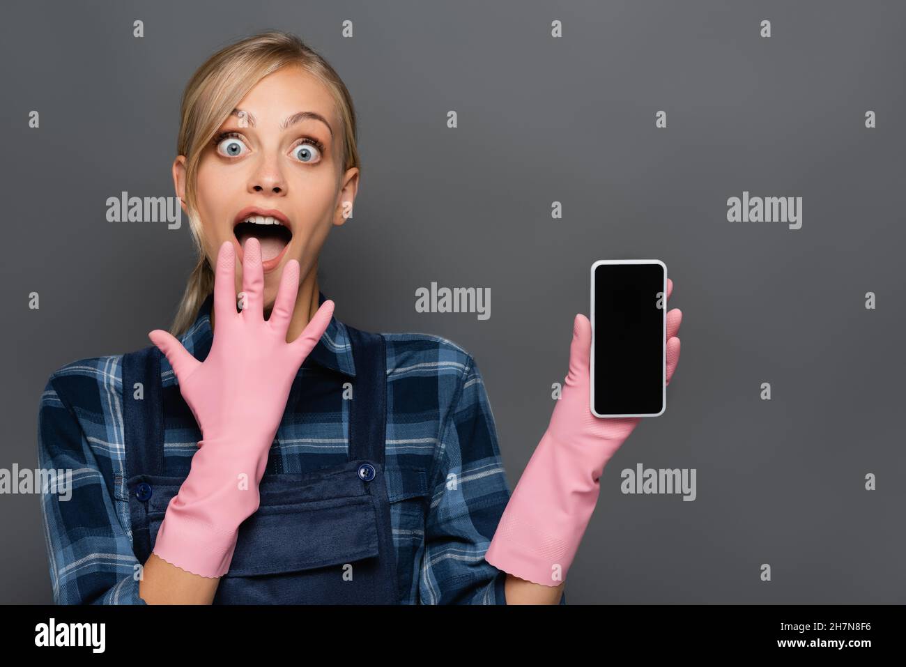 Astonished blonde plumber in rubber gloves holding cellphone with blank ...