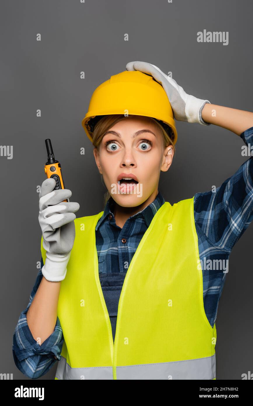 Scared builder in hardhat and safety vest holding walkie talkie and looking at camera isolated ...