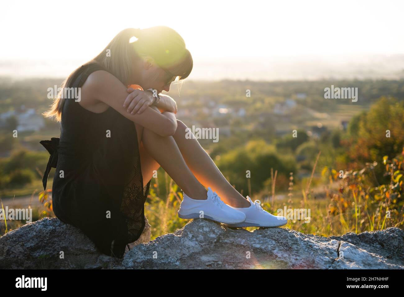 Pretty sad woman in black short summer dress sitting on a rock thinking ...