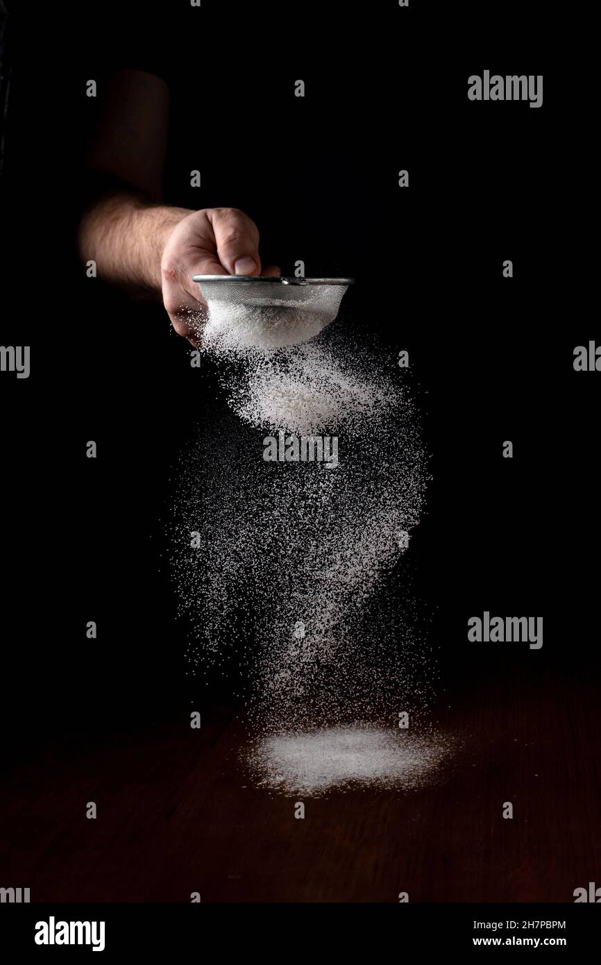 Flour flies through a sieve on a black background. A man sifts flour ...