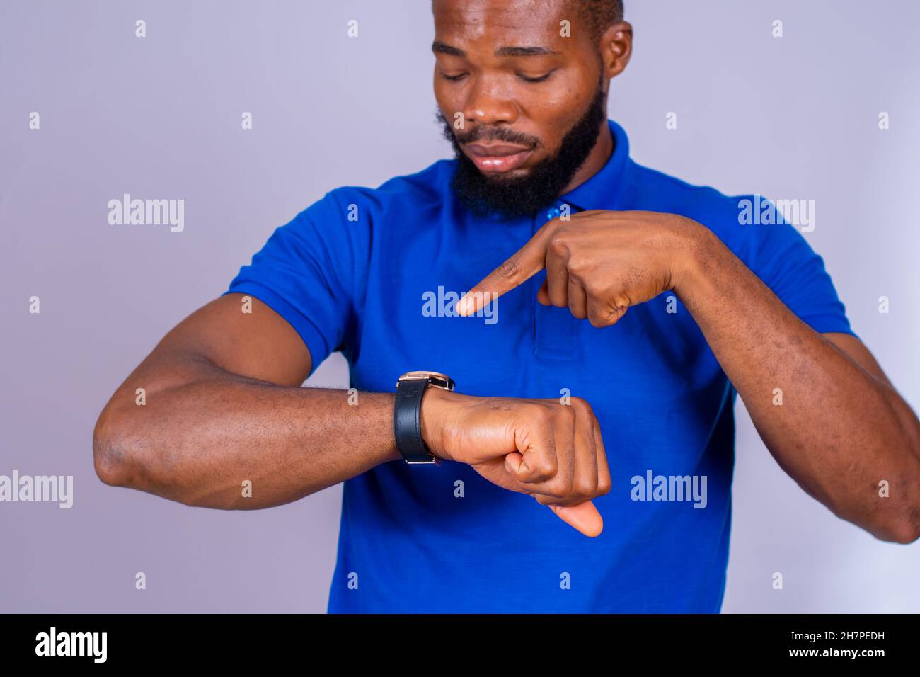 Young African male pointing at his watch standing on a pale purple ...