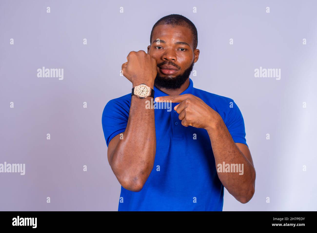 Young African male pointing at his watch standing on a pale purple ...