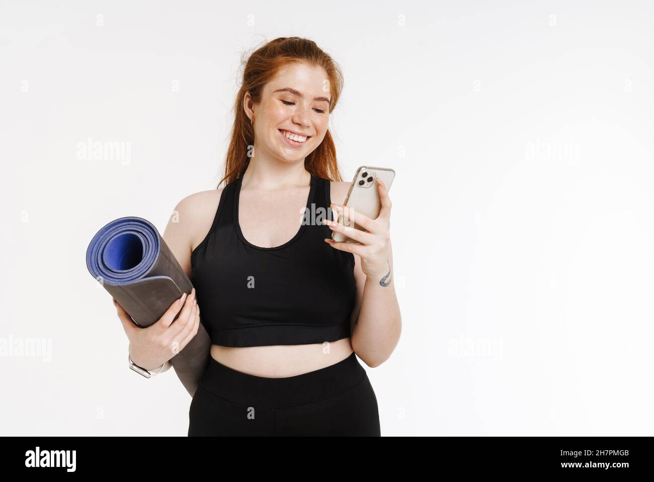 Young ginger woman using cellphone while posing with fitness mat ...