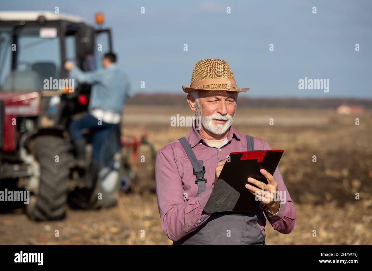 Senior farmer writing notes in notepad in field with tractor in ...