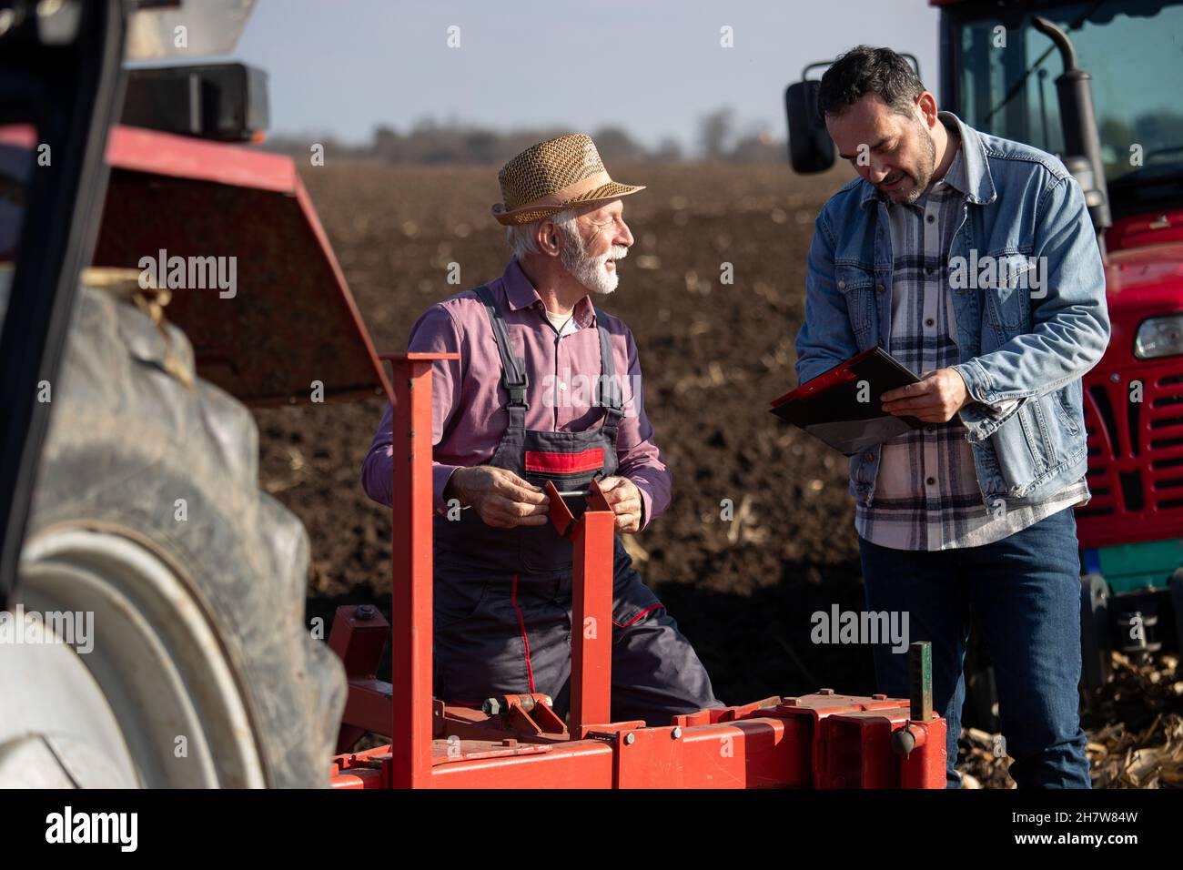 Senior farmer working on equipment of tractor while younger man writing ...