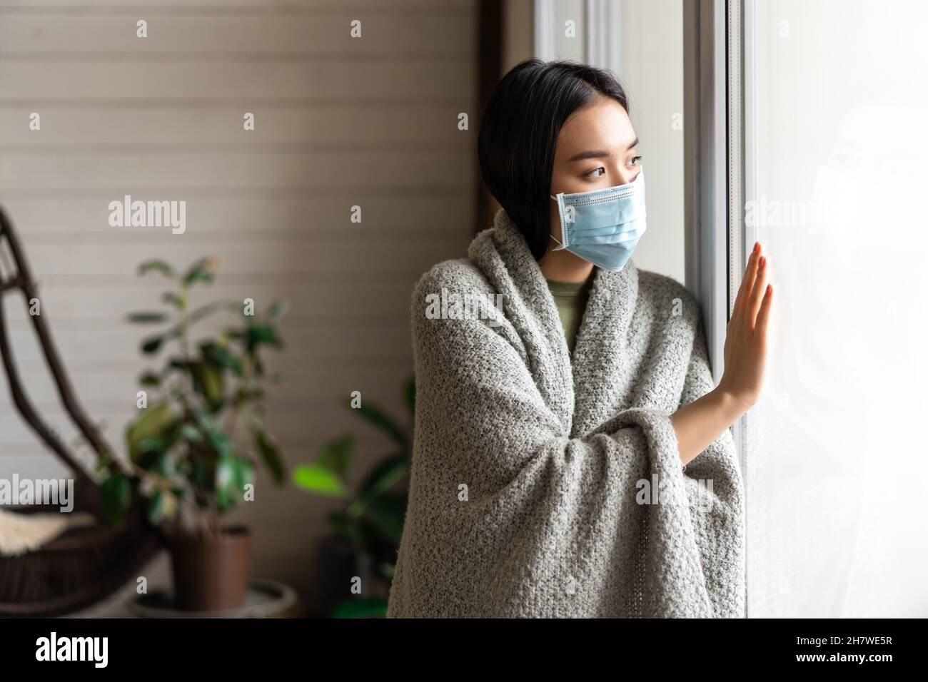 Sick asian girl in medical face mask standing by the window and ...