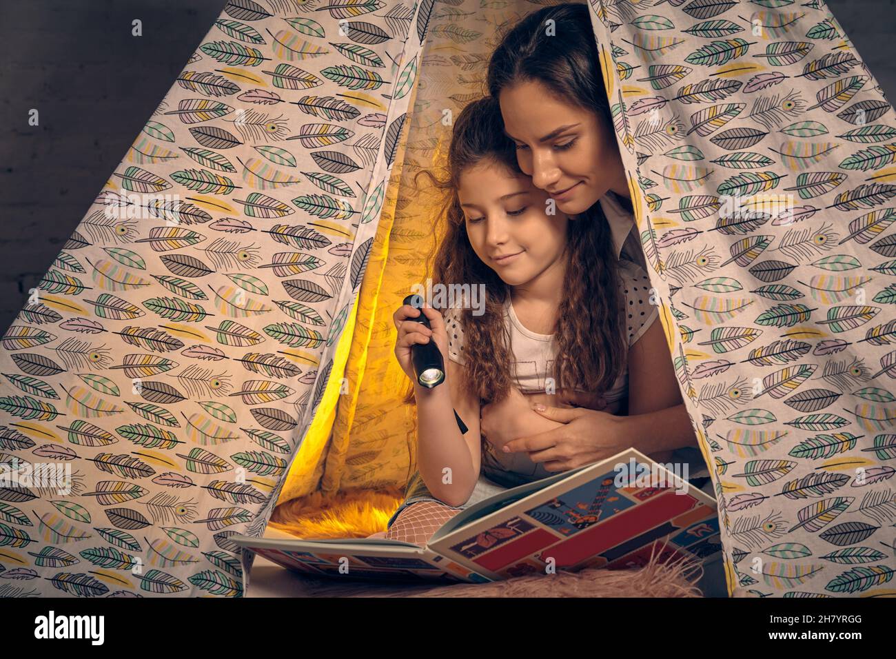 Mother and daughter are sitting in a teepee tent, reading stories with ...