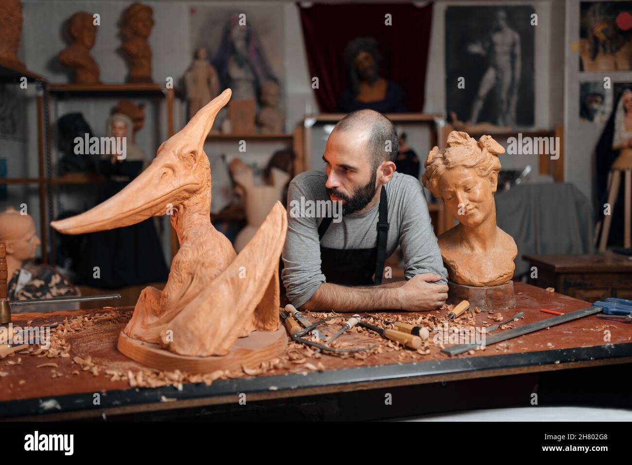 Pensive bearded male woodworker in apron standing at table with carved ...