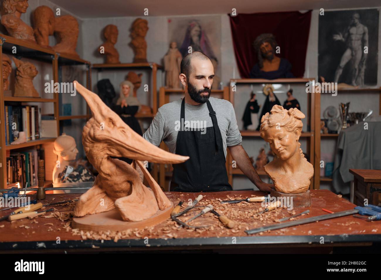 Pensive bearded male woodworker in apron standing at table with carved ...