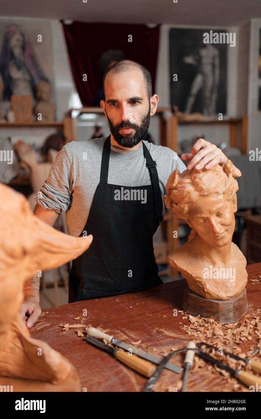 Pensive bearded male woodworker in apron standing at table with carved ...