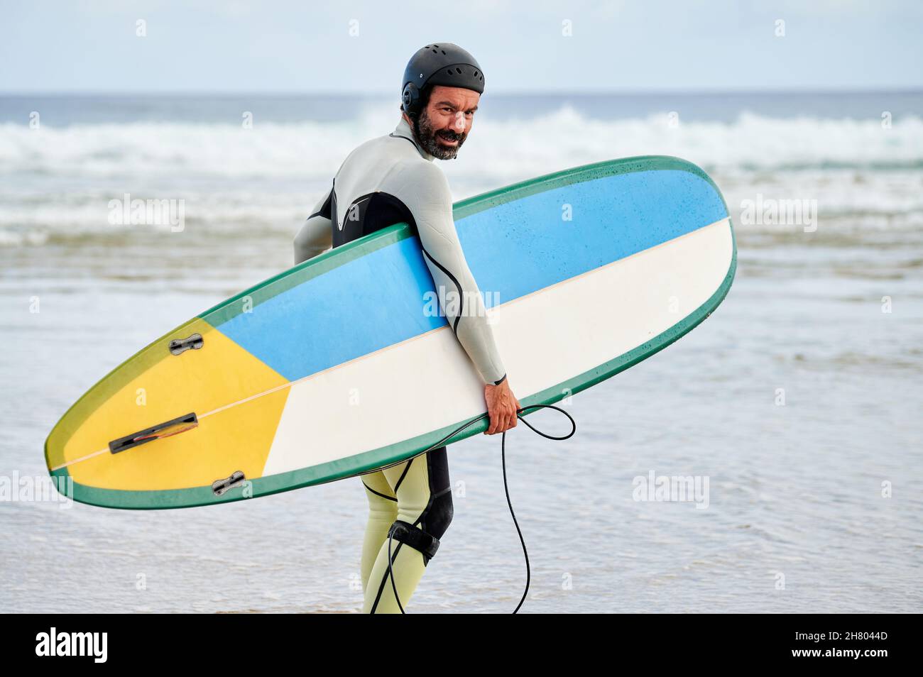 Back view of male surfer in wetsuit and helmet carrying surfboard ...